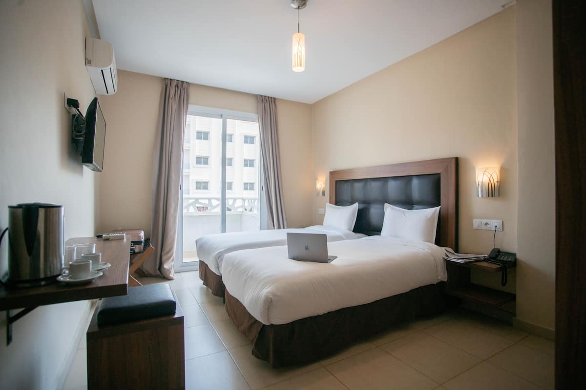 Quarto de hotel com duas camas, uma mesa com um laptop, uma cafeteira e vista para o exterior.