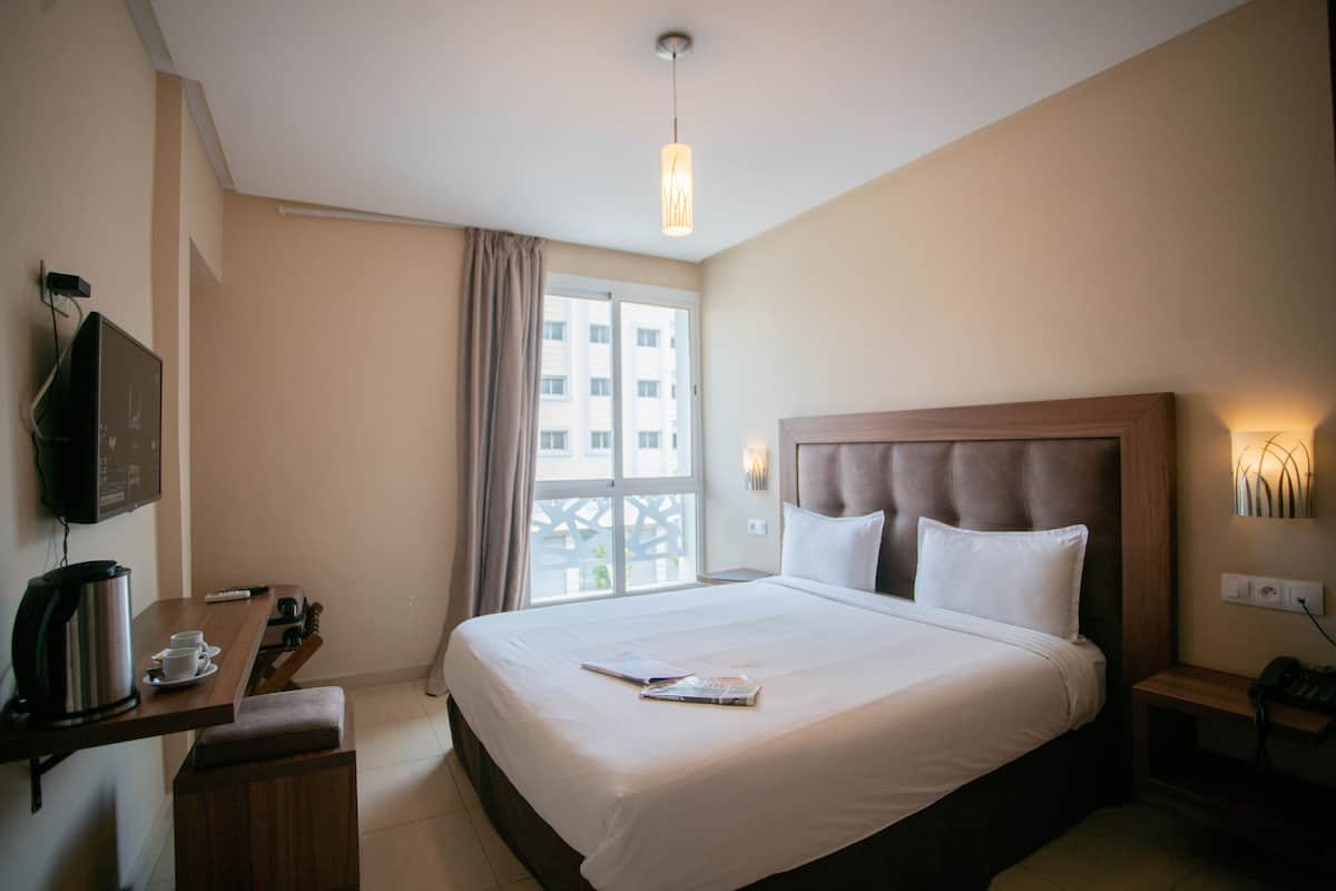 Quarto de hotel com cama, escrivaninha com cafeteira, televisão e janela com cortinas.