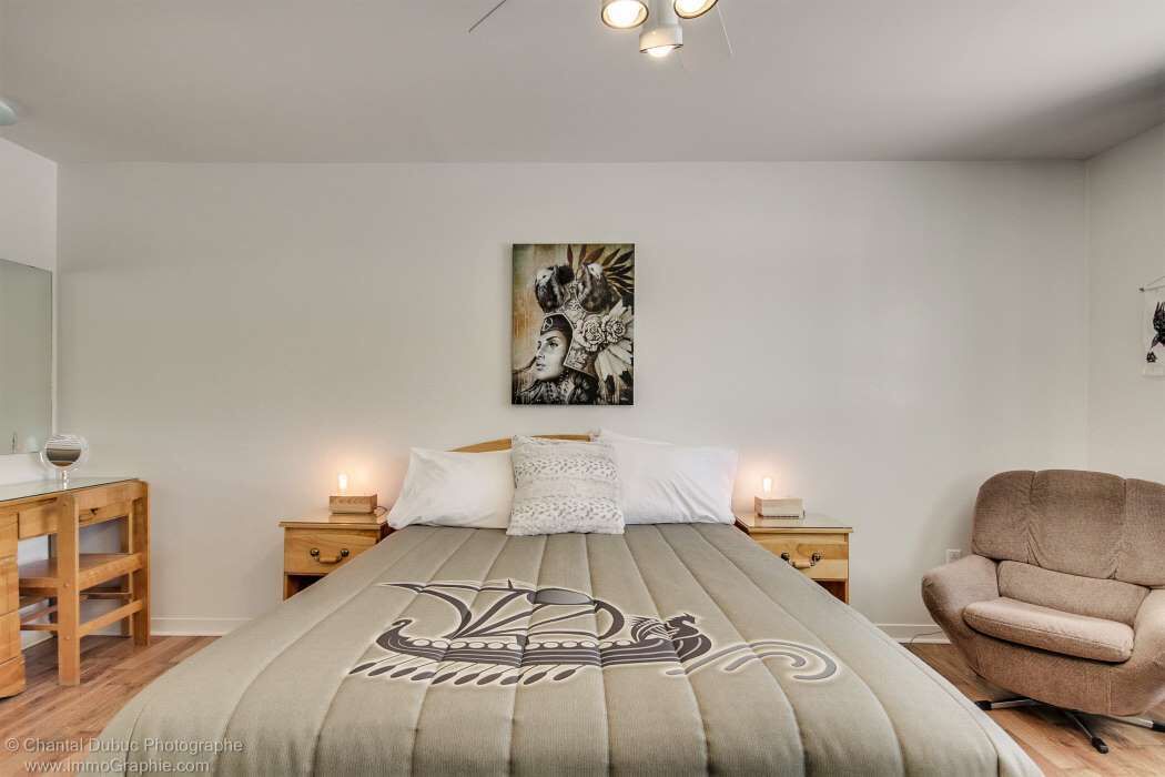 Quarto com cama, mesas de cabeceira, uma cadeira, um quadro na parede e uma mesinha de madeira.
