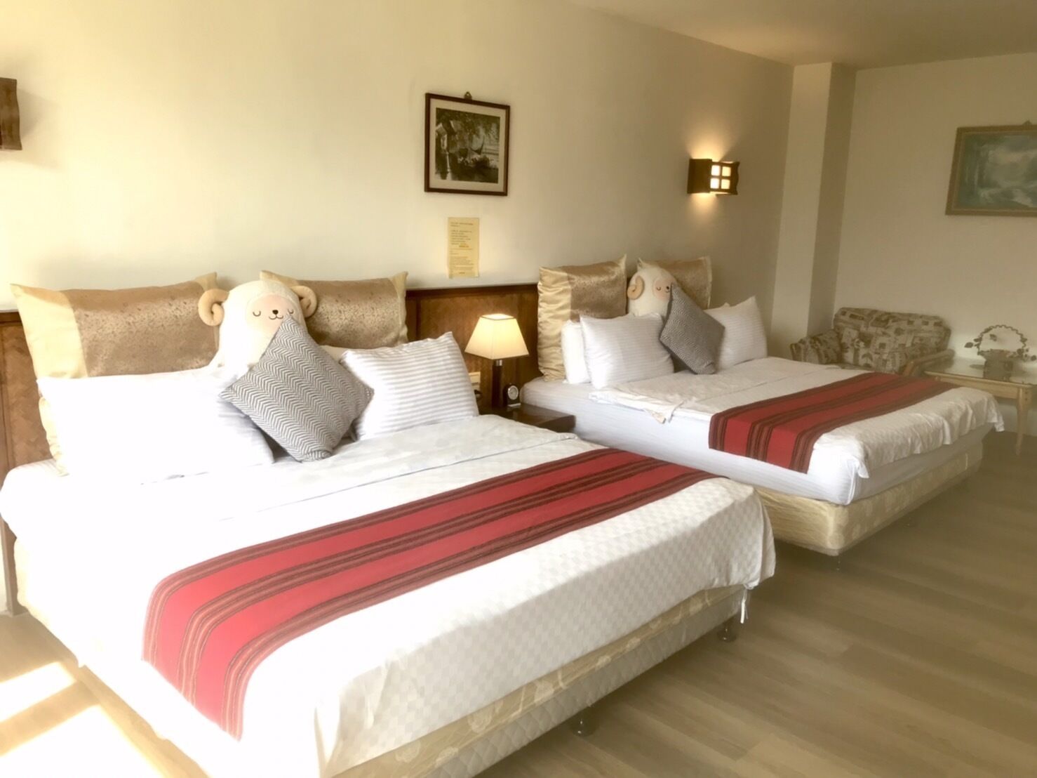 Quarto de hotel com duas camas, cada uma com um cobertor vermelho e branco, uma mesinha de cabeceira com uma luminária e um quadro na parede.