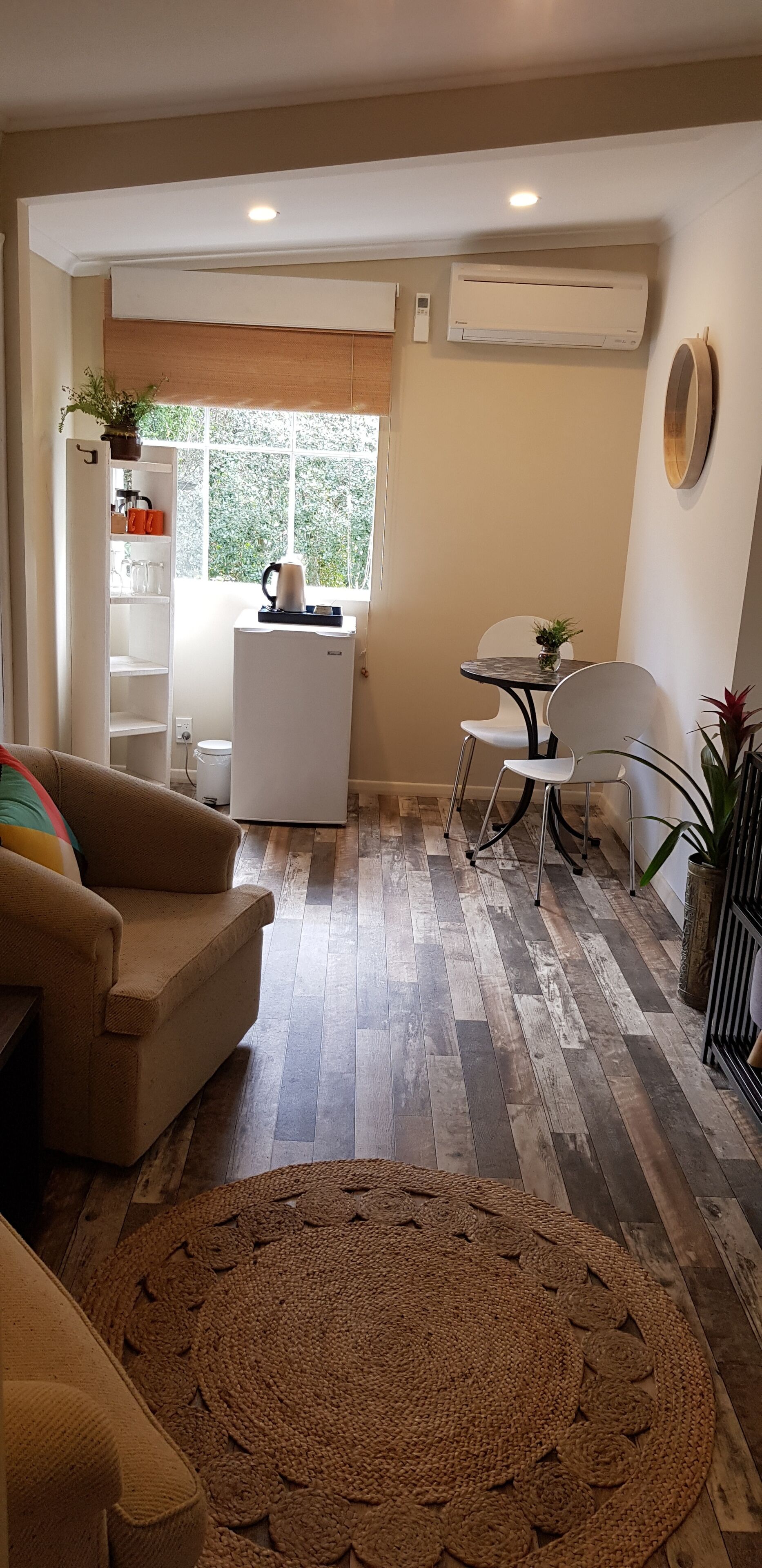 Um espaço de estar compacto com sofá, mesa de jantar com cadeiras, cozinha com geladeira e janela que proporciona iluminação natural.