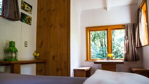 Quarto com cama, paredes de madeira, janela com vista para vegetação, pequena mesa com um vaso de flores e um lampião verde.