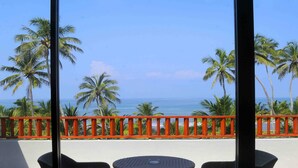 Varanda com duas cadeiras e uma mesa, com vista para uma praia com palmeiras e o oceano.