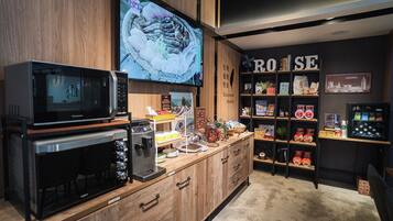 Cozinha compacta com micro-ondas, cafeteira e televisão exibindo uma cena de comida. Prateleiras com lanches e um aviso com a inscrição “ROSE” são visíveis.
