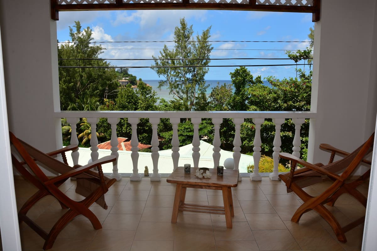Varanda com mesa de madeira e duas cadeiras de madeira, guarda-corpo branco, vista para árvores e um edifício na água.