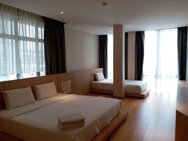 Quarto de hotel com uma cama grande, uma cama menor e uma cama-sofá. Há cortinas nas janelas e o chão é de madeira.