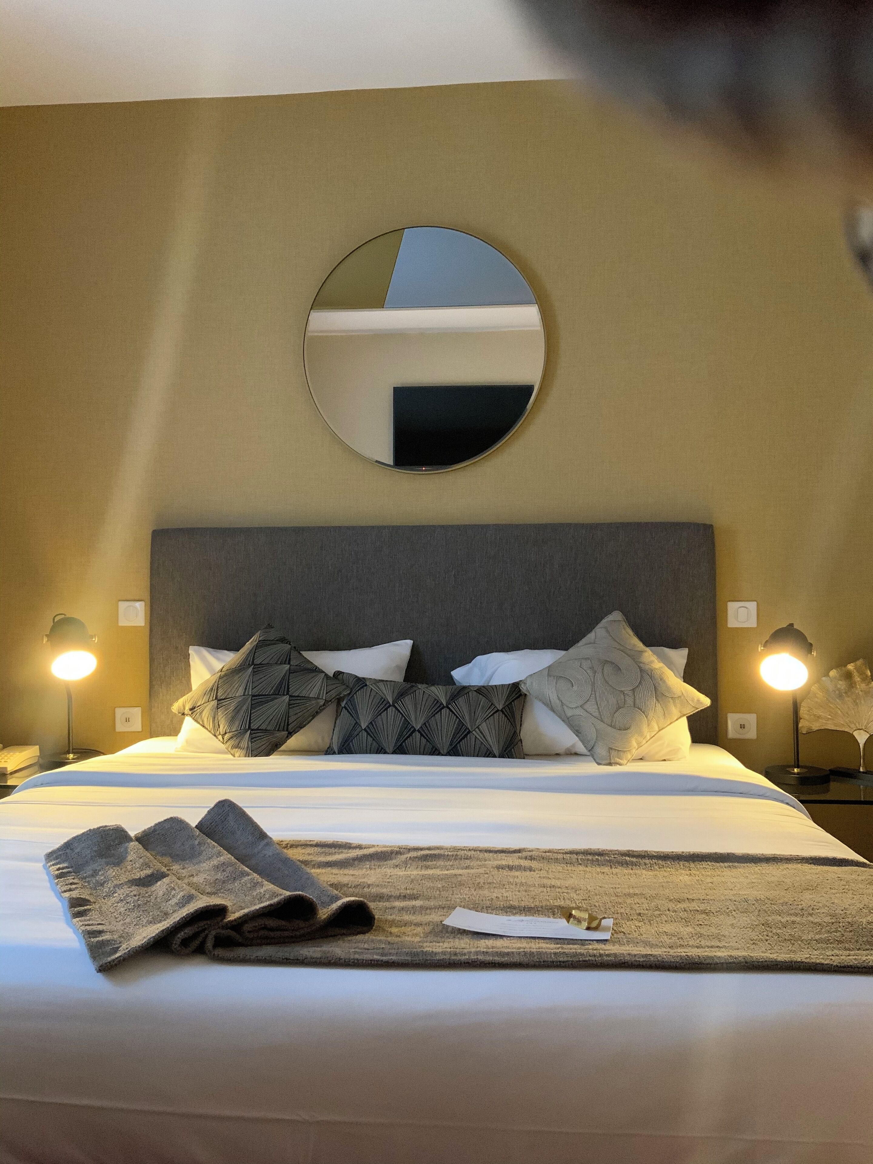 Uma cama bem arrumada com almofadas estampadas, um espelho redondo na parede e duas luminárias de cabeceira.