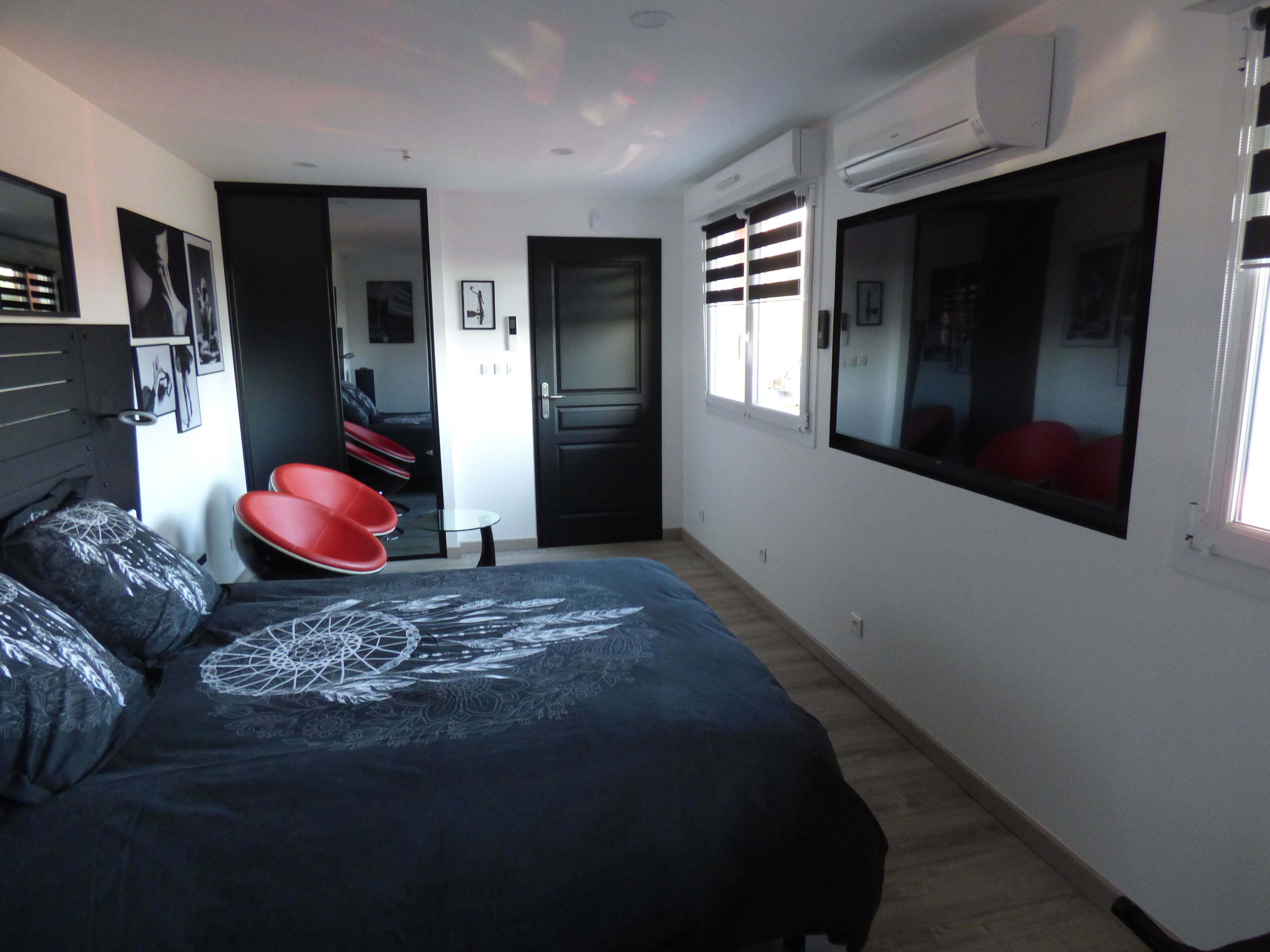Um quarto com cama, uma cadeira vermelha, uma mesa pequena e uma janela grande.