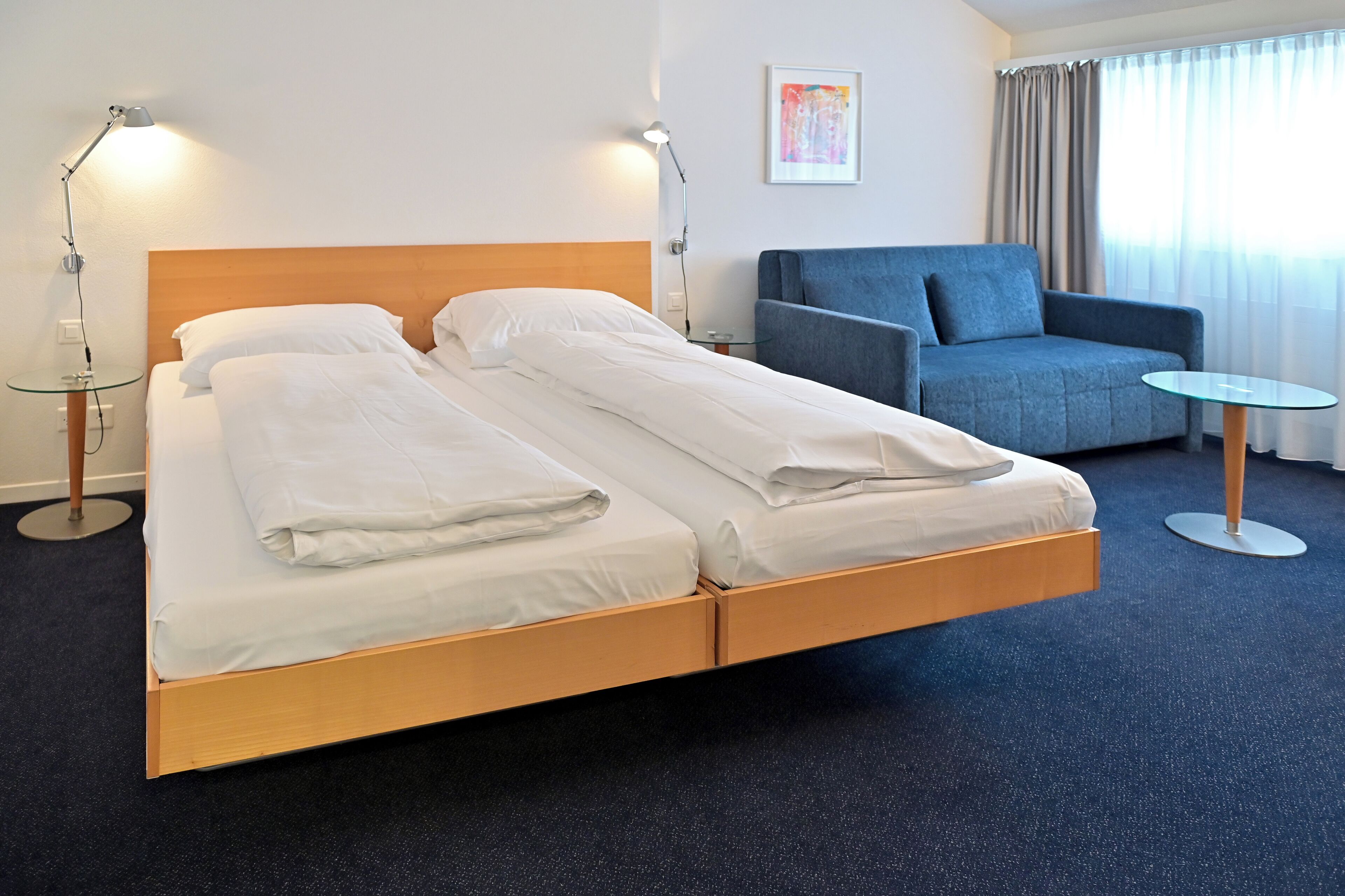 Quarto de hotel com cama de casal, sofá azul, mesa redonda pequena e duas luminárias de cabeceira.