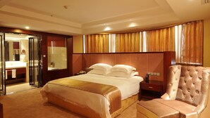 Quarto de hotel com uma cama grande, área de estar, penteadeira com espelho e janela com cortinas.