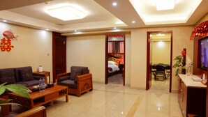 Quarto de hotel com sofá, poltrona, mesa de centro de madeira e televisão. Há uma porta que dá para um quarto e uma área de refeições.