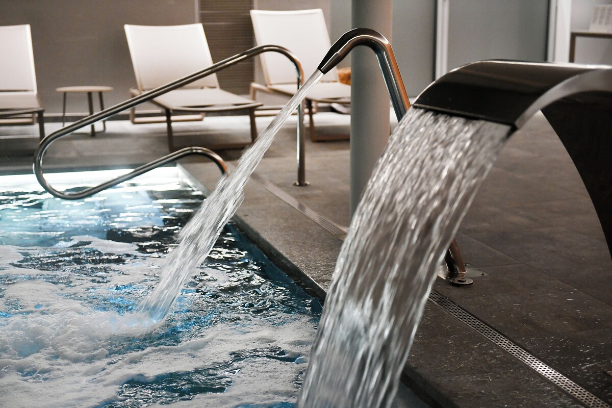 Uma piscina com água escorrendo por uma borda de metal, rodeada por espreguiçadeiras e um piso de azulejos.