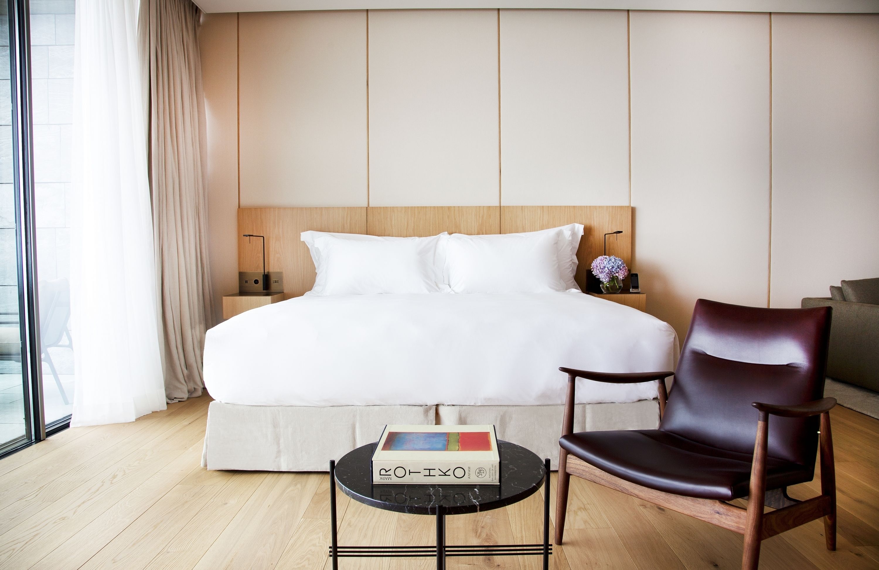 Quarto de hotel com uma cama grande, cabeceira de madeira, mesa redonda preta com um livro, poltrona de couro marrom e uma janela ampla com cortinas.