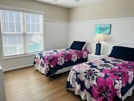 Quarto com duas camas, uma mesinha de cabeceira com abajur, janela com persianas e um quadro na parede.