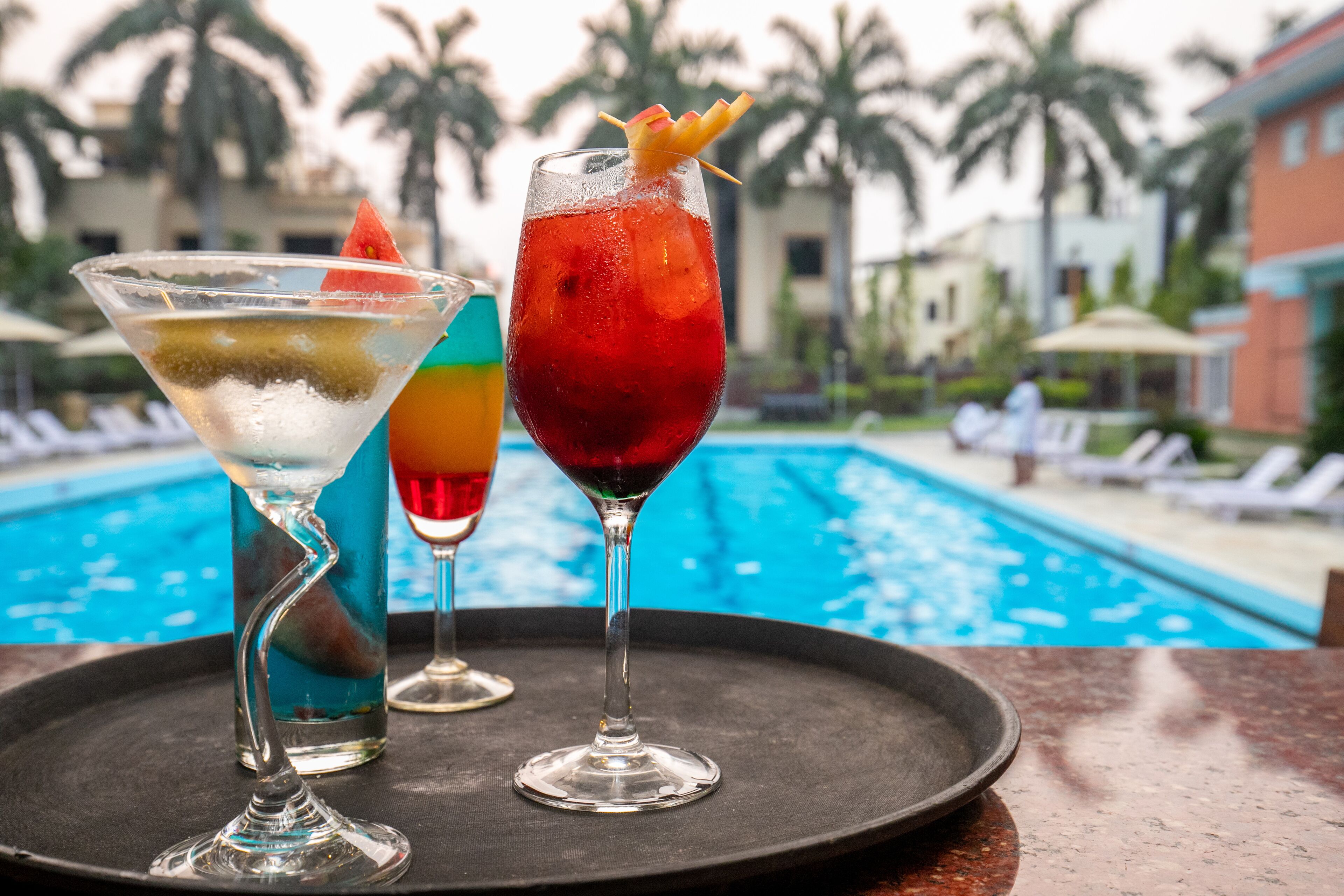 Dois cocktails numa bandeja perto de uma piscina, com palmeiras e um edifício ao fundo.