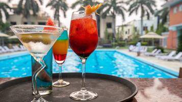 Dois cocktails numa bandeja perto de uma piscina, com palmeiras e um edifício ao fundo.