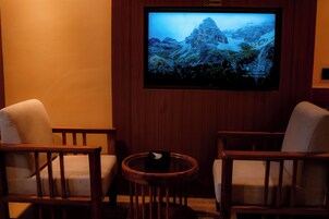 Quarto com duas cadeiras de madeira, uma mesa redonda pequena e uma televisão fixada em parede de madeira exibindo uma paisagem de montanhas.