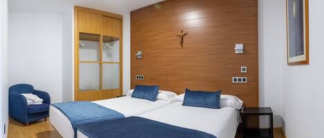 Um quarto com duas camas, uma cabeceira de madeira, uma cadeira azul, uma mesinha de cabeceira e um quadro na parede.