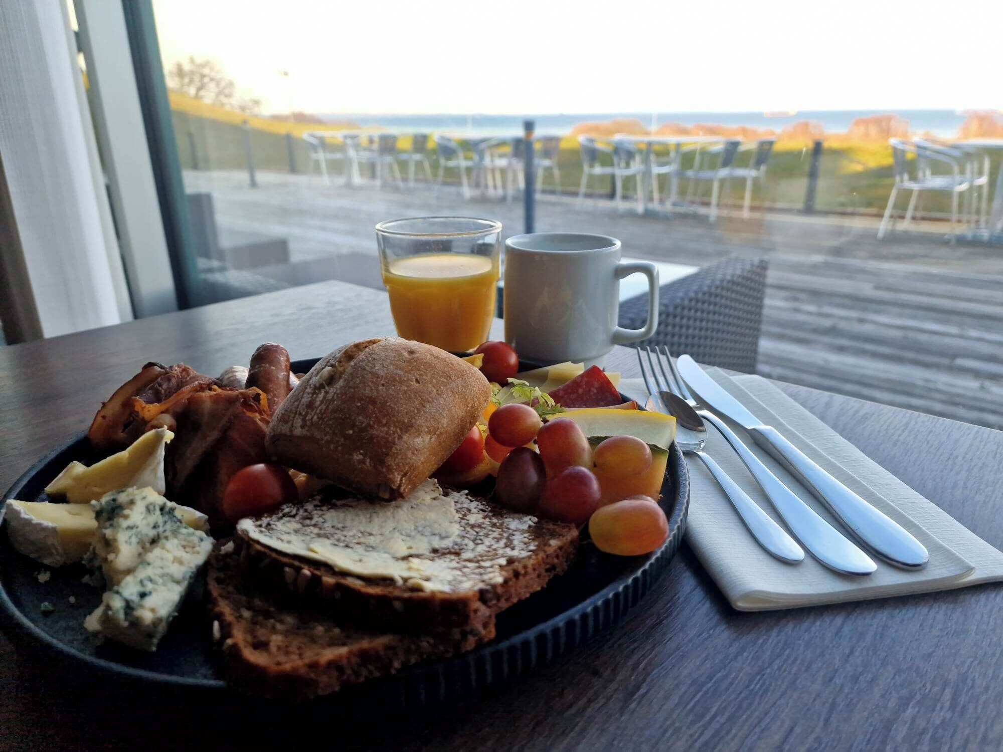 Prato de pequeno-almoço com pão, queijo, fiambre e fruta, um copo de sumo de laranja e uma chávena sobre uma mesa com vista para mesas ao ar livre.