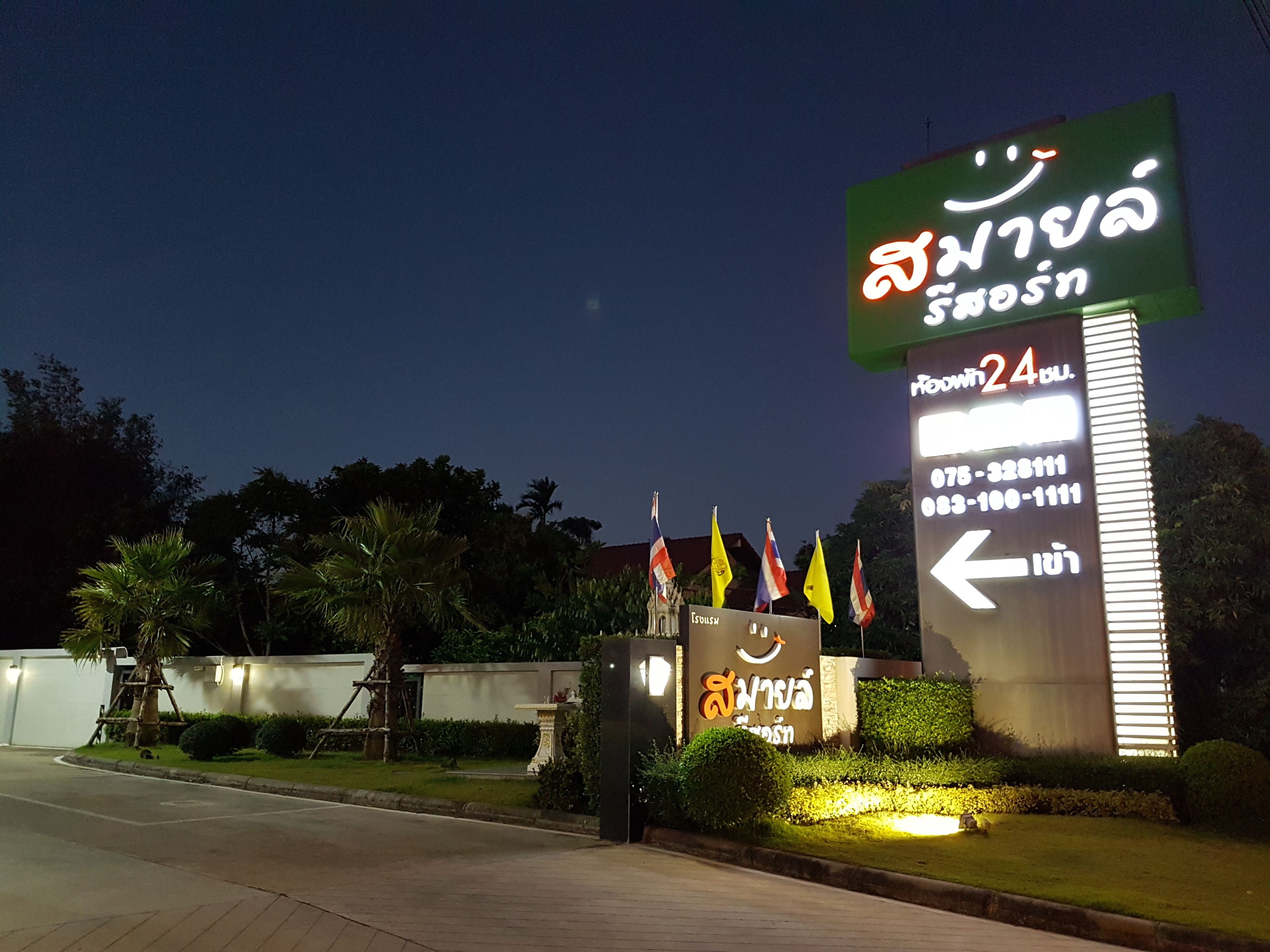 Entrada de hotel com placa indicando serviço 24 horas, números de contato e setas de direção.