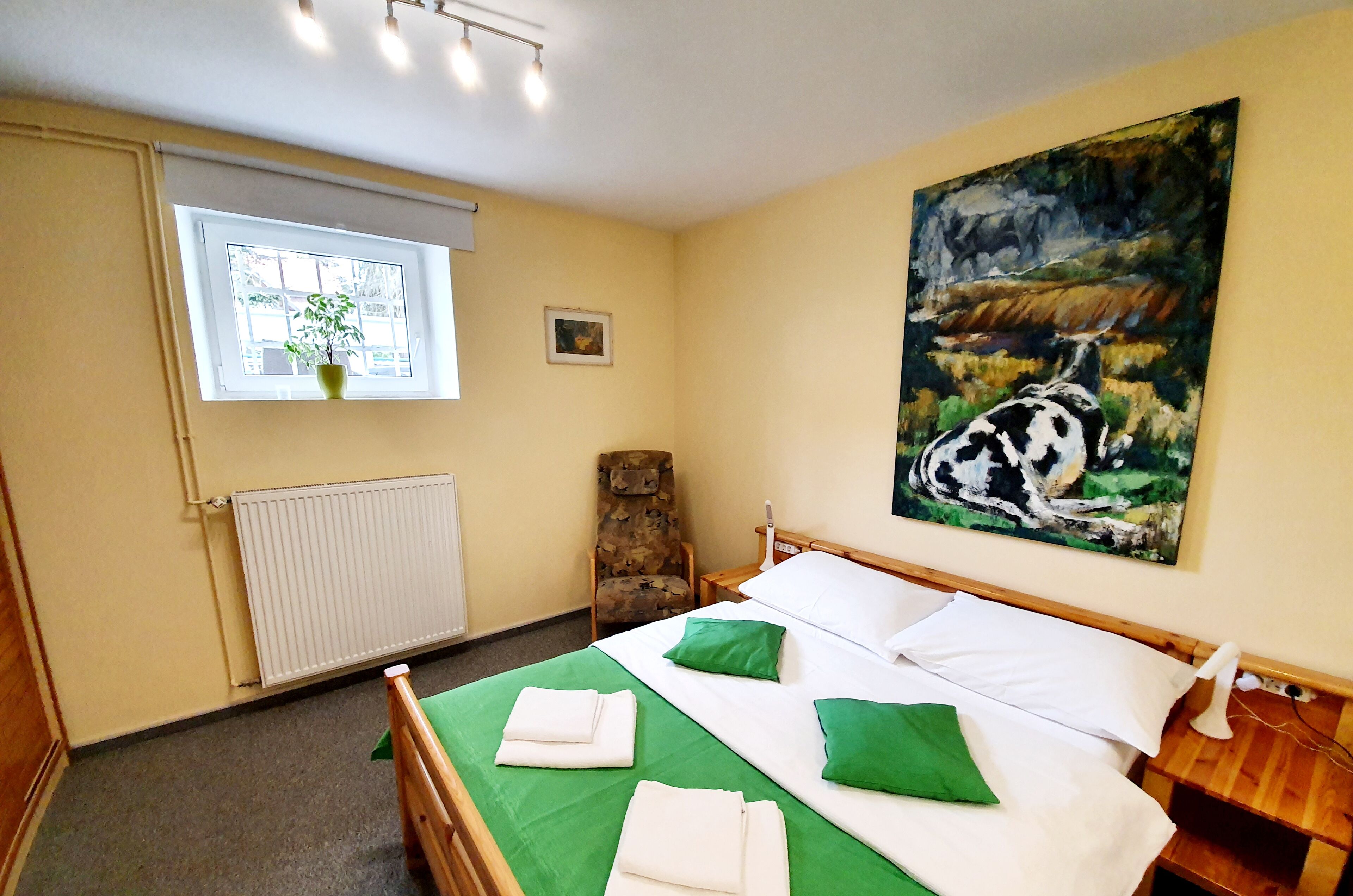 Quarto de hotel com cama, uma cadeira, radiador, janela com planta e pintura na parede.