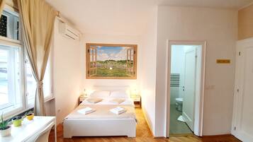 Quarto com cama, luminárias de cabeceira, escrivaninha, planta em vaso e pintura de paisagem.