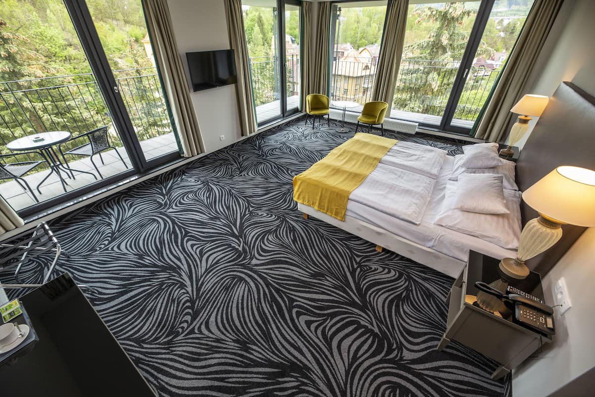 Quarto de hotel com carpete estampado em zebra, uma cama com lençóis brancos, duas cadeiras amarelas, uma mesa pequena e uma varanda com vista para vegetação e edifícios.
