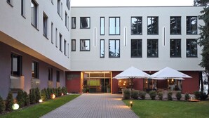 Entrada de hotel com calçada, área de estar ao ar livre com guarda-sóis e gramado bem cuidado.