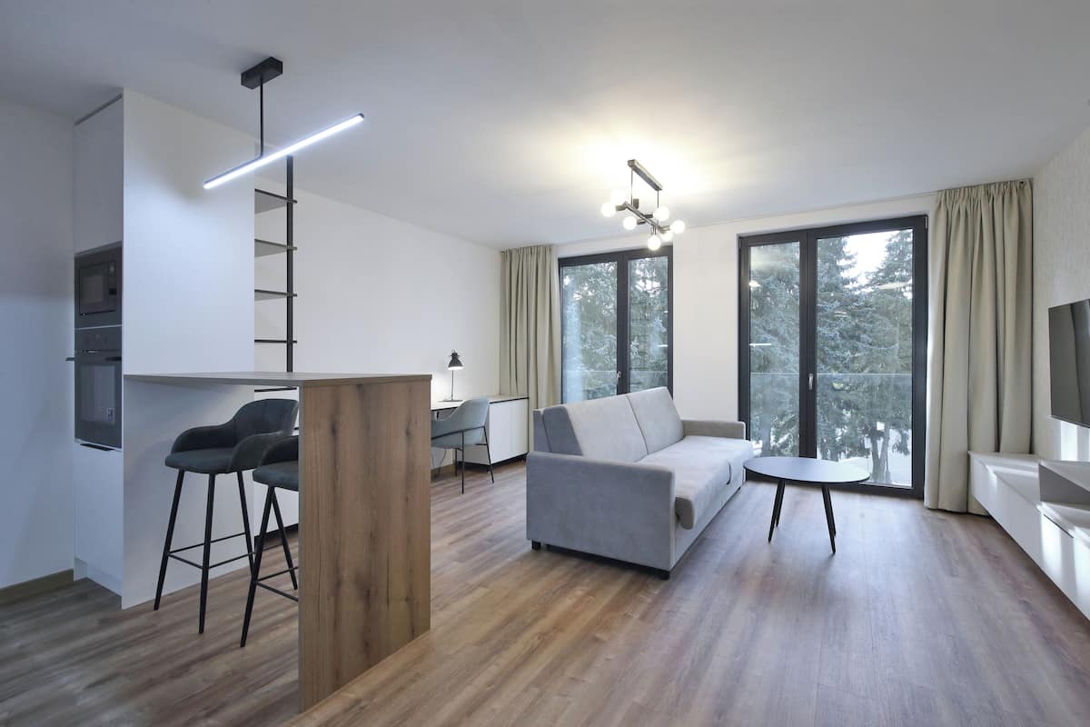 Uma sala de estar moderna com sofá, mesa de jantar e TV. Há uma área de cozinha com micro-ondas e bancada de madeira. A sala possui janelas amplas com cortinas.