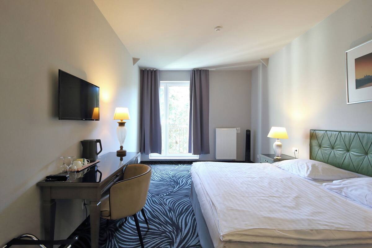 Quarto de hotel com cama, escrivaninha, cadeira, televisão e janela com cortinas.