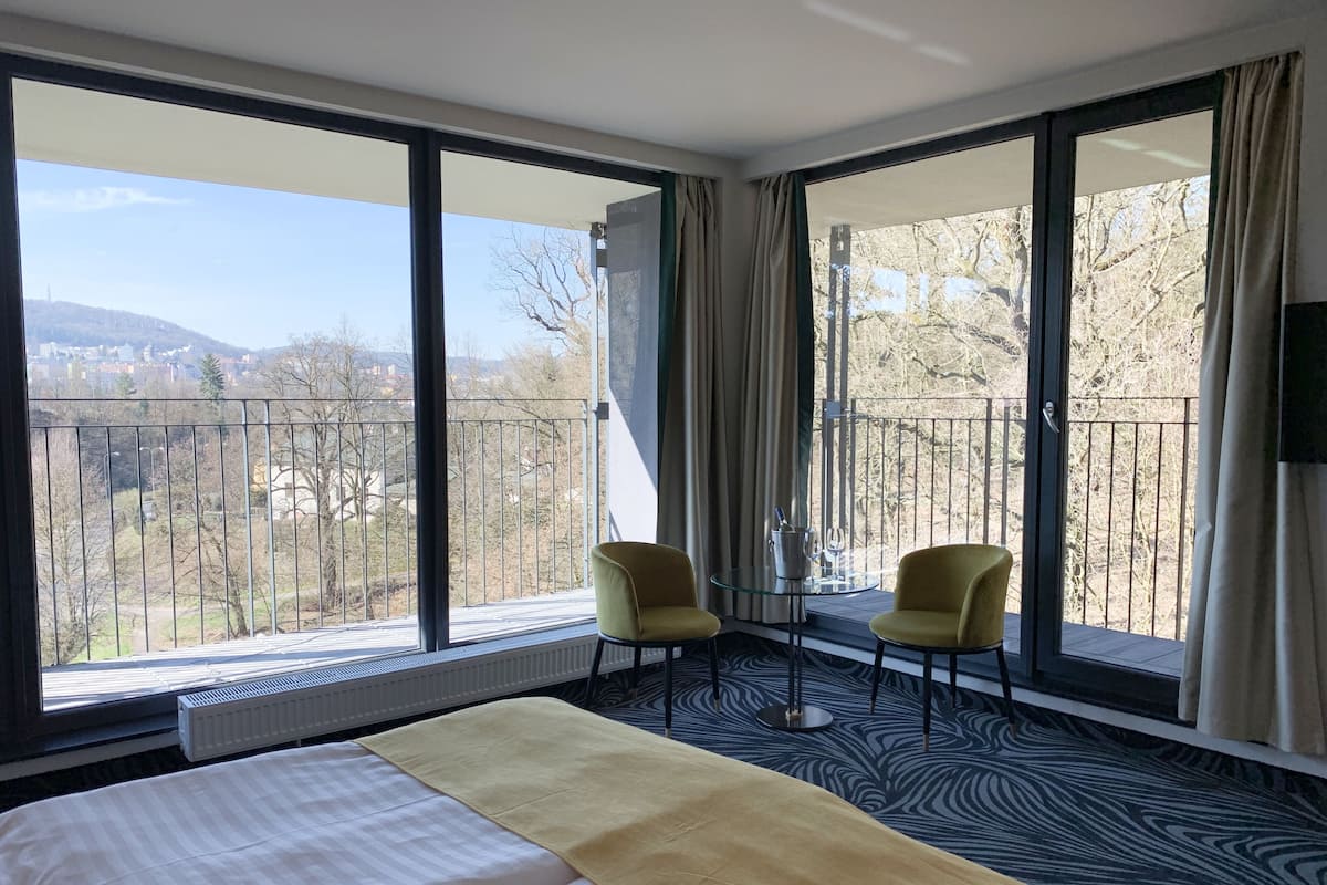 Quarto de hotel com uma cama grande, duas cadeiras, uma mesa pequena e uma varanda com vista para a cidade e para as árvores.