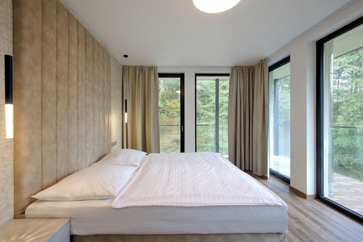 Quarto moderno com cama grande, cabeceira de madeira e janelas amplas com vista para árvores.