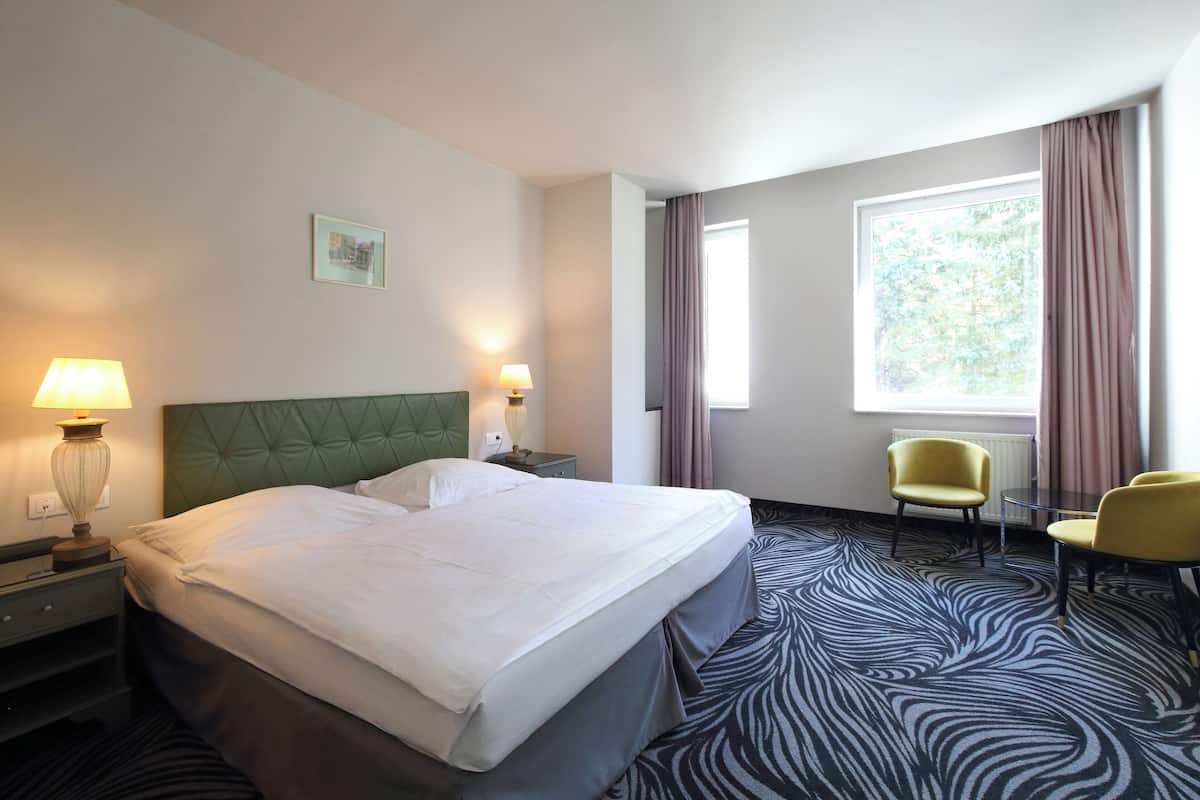 Quarto de hotel com uma cama grande, duas luminárias laterais, cabeceira verde, tapete com padrão de zebra, janela com cortinas e uma pequena mesa com uma cadeira.