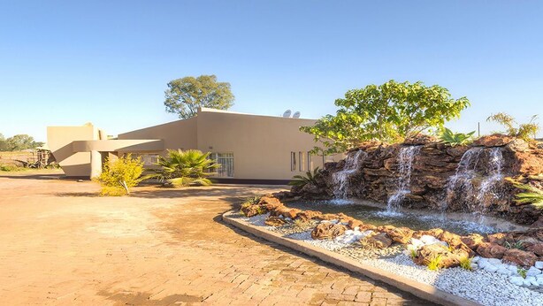 Área ajardinada com elemento aquático, incluindo uma pequena cascata e vegetação ao redor, em frente a um edifício moderno com telhado plano e antenas parabólicas.