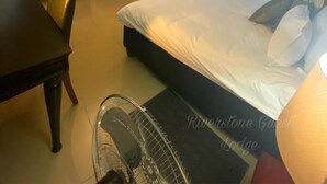 Quarto de hotel com uma cama, uma escrivaninha, duas cadeiras, uma televisão e um ventilador.