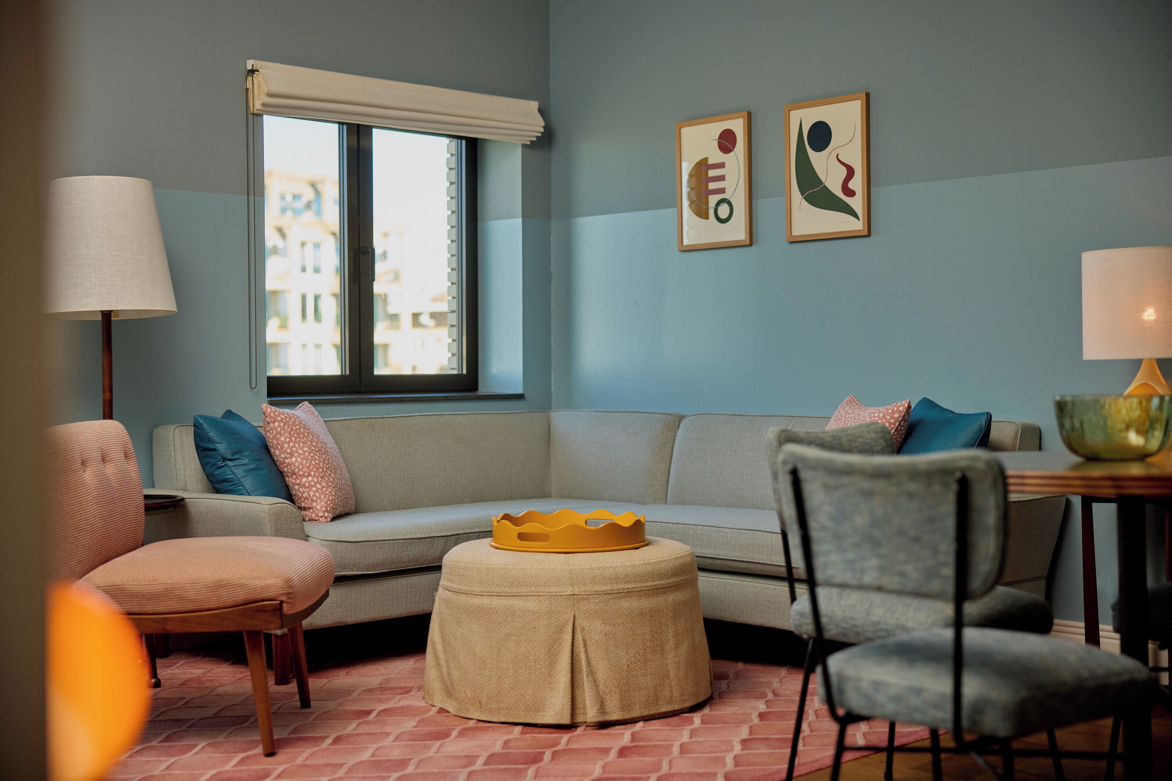Uma sala de estar com sofá, banqueta, cadeiras, mesa com abajur, janela com persianas e duas obras de arte nas paredes.