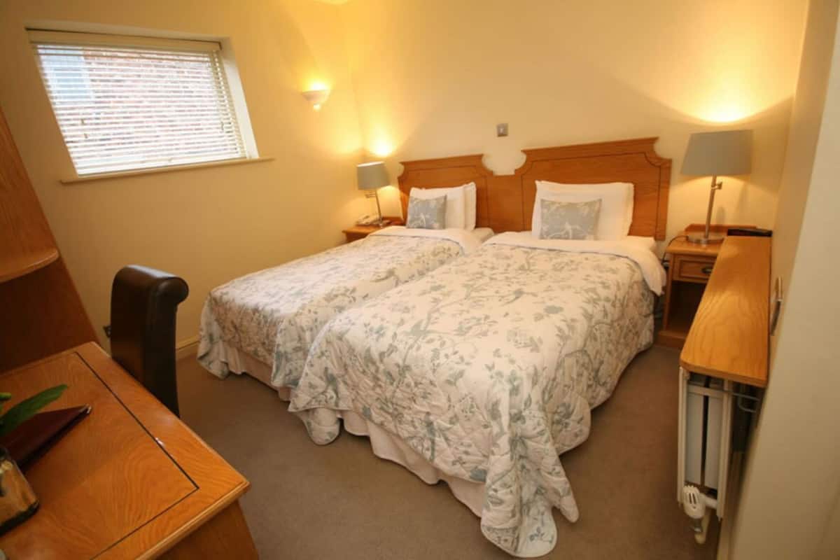 Uma cama de casal com roupa de cama floral, luminárias de cabeceira, uma cabeceira de madeira, uma mesinha com uma planta e um radiador.