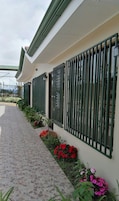 Uma trilha conduz a um edifício com piso de azulejos, guarda-corpos de metal verde e canteiros com flores vermelhas e cor-de-rosa.