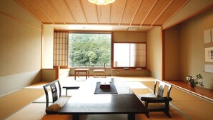 Um quarto japonês tradicional com tatames, uma mesa baixa, cadeiras e vista para uma floresta através de grandes janelas.