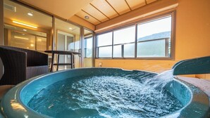 Uma banheira de hidromassagem com água a correr, uma cadeira de vime e vista para um corpo de água através de grandes janelas.