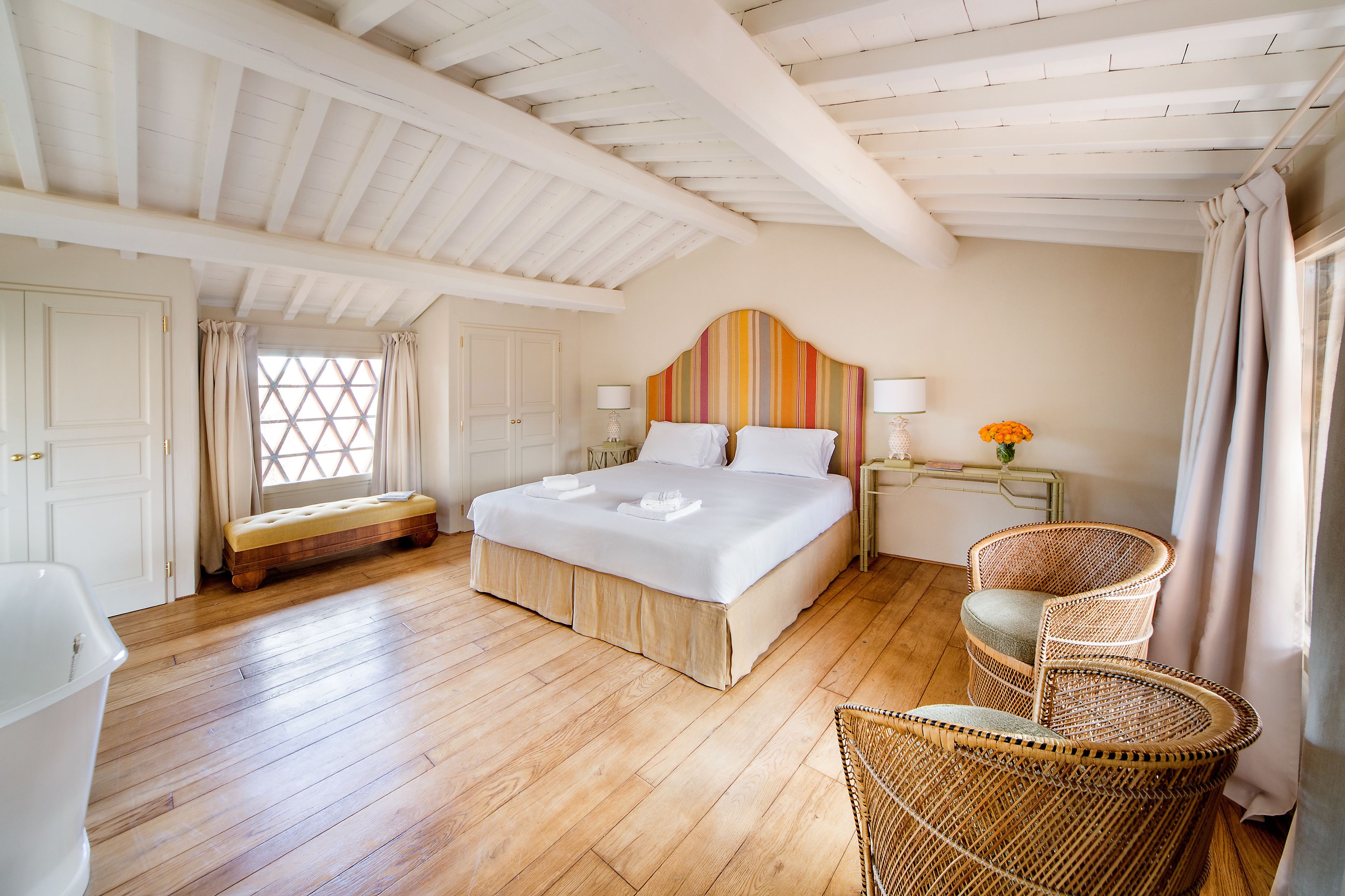 Um quarto espaçoso com uma cama grande, piso de madeira, um banco, cadeiras de vime e uma janela com cortinas.