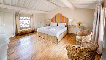 Um quarto espaçoso com uma cama grande, piso de madeira, um banco, cadeiras de vime e uma janela com cortinas.