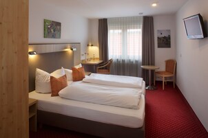 Quarto de hotel com cama, secretária, cadeira, televisão e janela com cortinas.