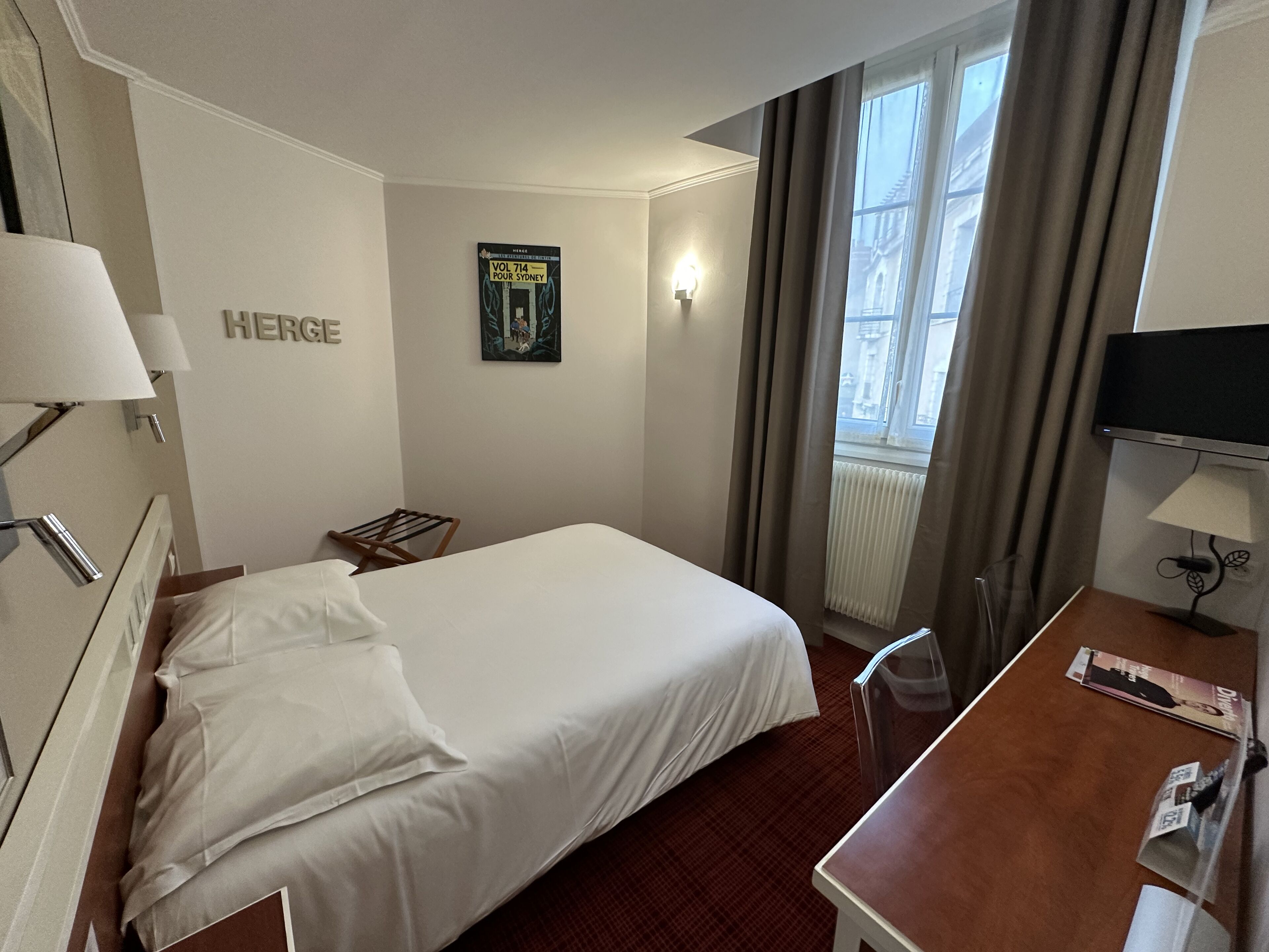 Quarto de hotel com cama, escrivaninha, cadeira, televisão, janela com cortinas, radiador e um cartaz na parede.