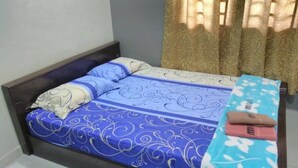 Uma cama com colcha azul e branca estampada, cabeceira de madeira e janela com cortinas padronizadas.