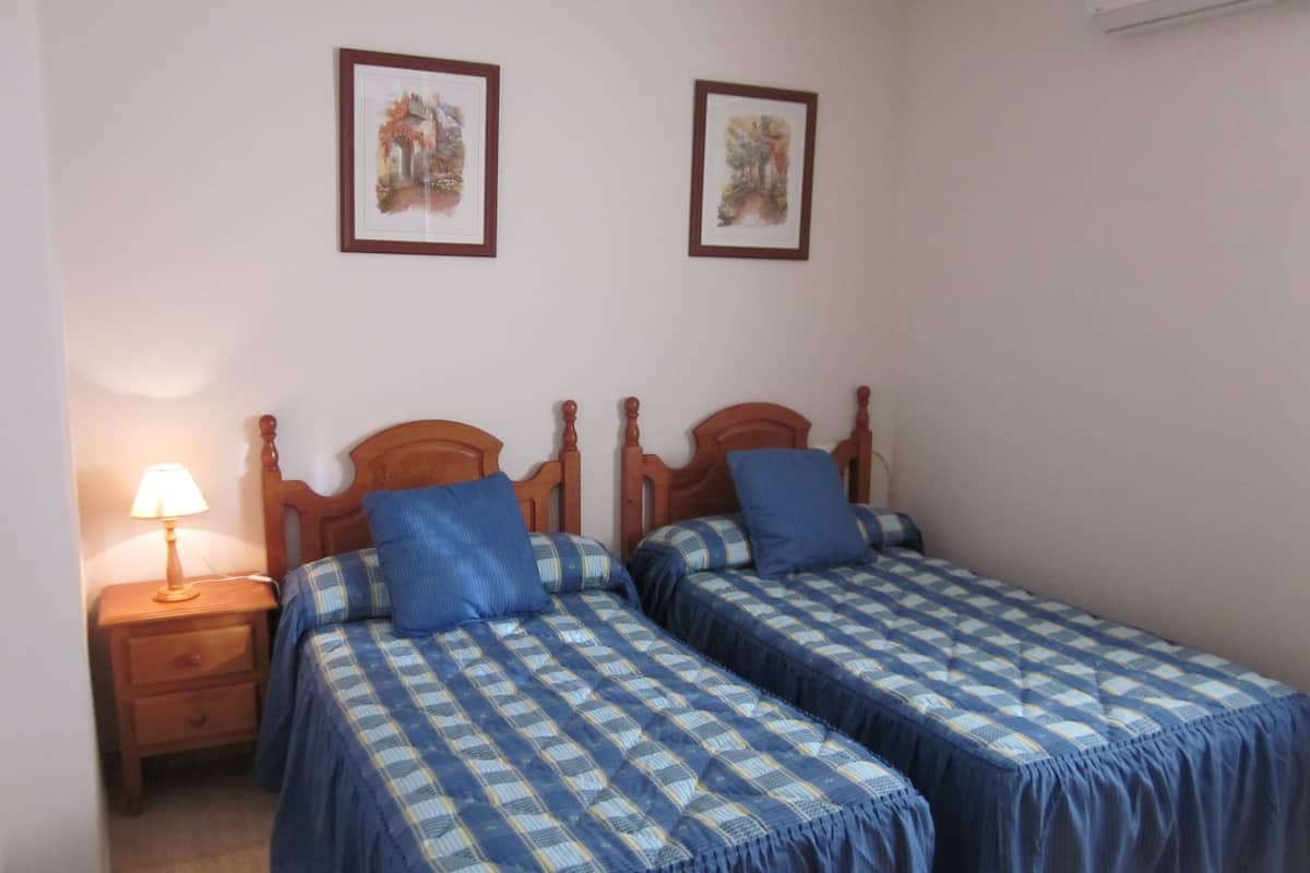 Quarto com duas camas de solteiro, cada uma com cabeceira de madeira e roupa de cama azul. Um criado-mudo com abajur fica do lado esquerdo. Duas imagens emolduradas pendem na parede acima das camas.