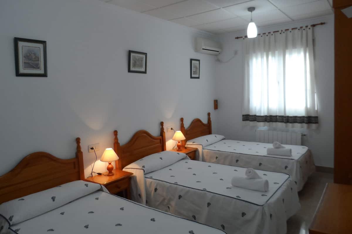 Quarto de hotel com três camas de solteiro, cada uma com cabeceira de madeira, luminária de cabeceira e colcha branca com padrão de folha. Há uma janela com cortinas brancas, um aparelho de ar condicionado na parede e quadros emoldurados.