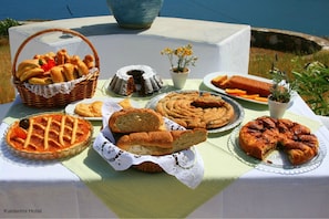 Uma mesa com diversos produtos de panificação, incluindo tortas, pão e um bolo bundt, disposta ao ar livre com vista para o mar.