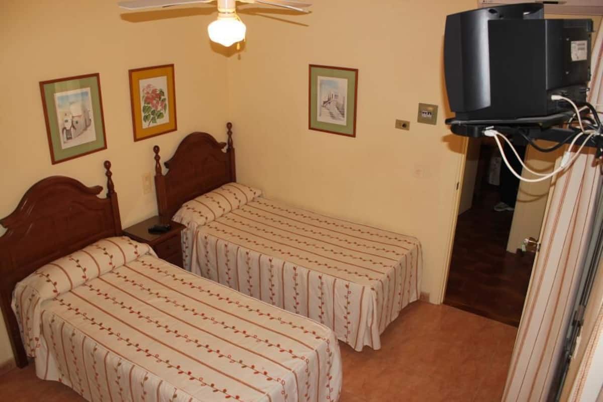 Quarto com duas camas de solteiro, um ventilador de teto e uma televisão fixada na parede.