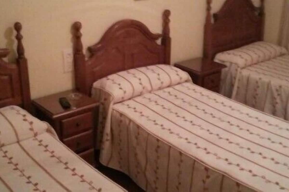 Quarto com três camas de madeira, cada uma com colcha estampada. Há mesas de cabeceira, com um controle remoto em uma delas.